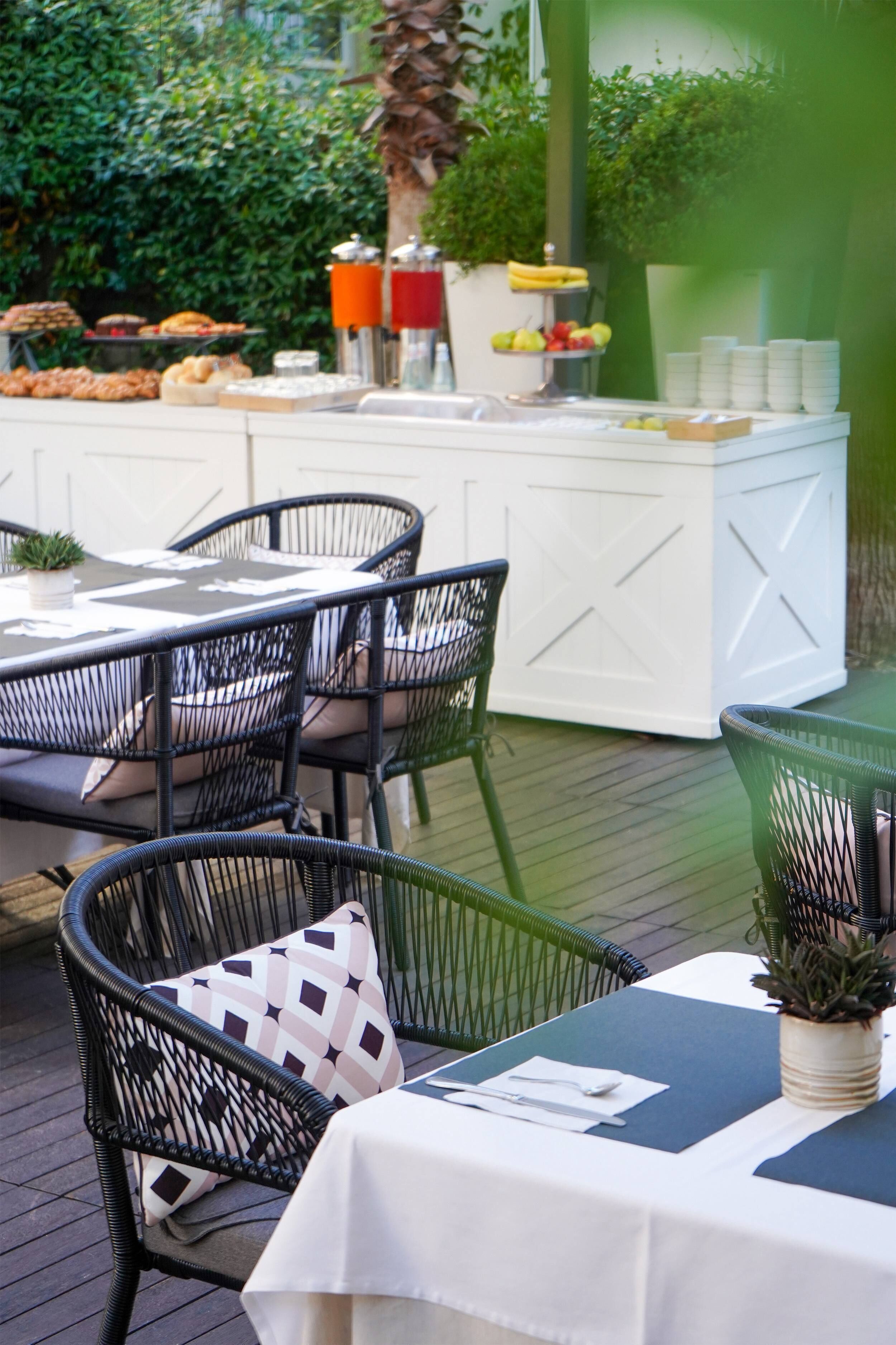 Área de refeições ao ar livre com um buffet branco, cadeiras de metal preto e uma mesa posta com toalhas brancas.