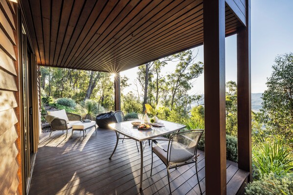 Um deck de madeira com mesa e cadeiras, rodeado por vegetação e vista para o mar.