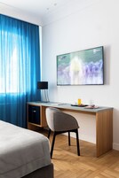 Quarto de hotel moderno com cama, escrivaninha, cadeira, televisão e janela com cortinas azuis.