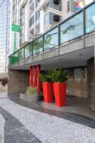 Entrada de hotel com design moderno, incluindo varanda de vidro, pilares vermelhos e plantas em vasos.