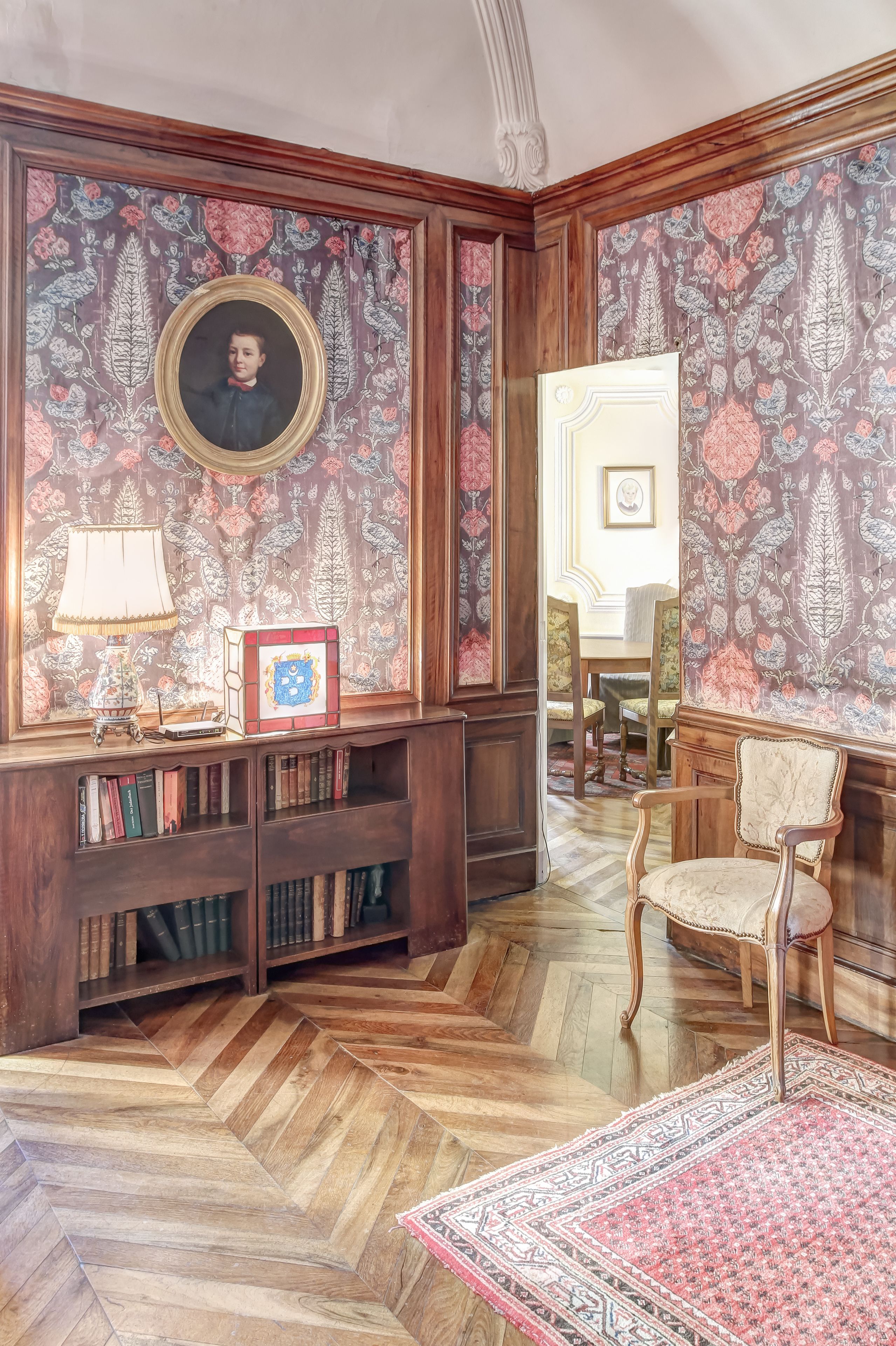 Quarto com piso de madeira em padrão herringbone, uma estante de madeira, um retrato emoldurado e papel de parede estampado.
