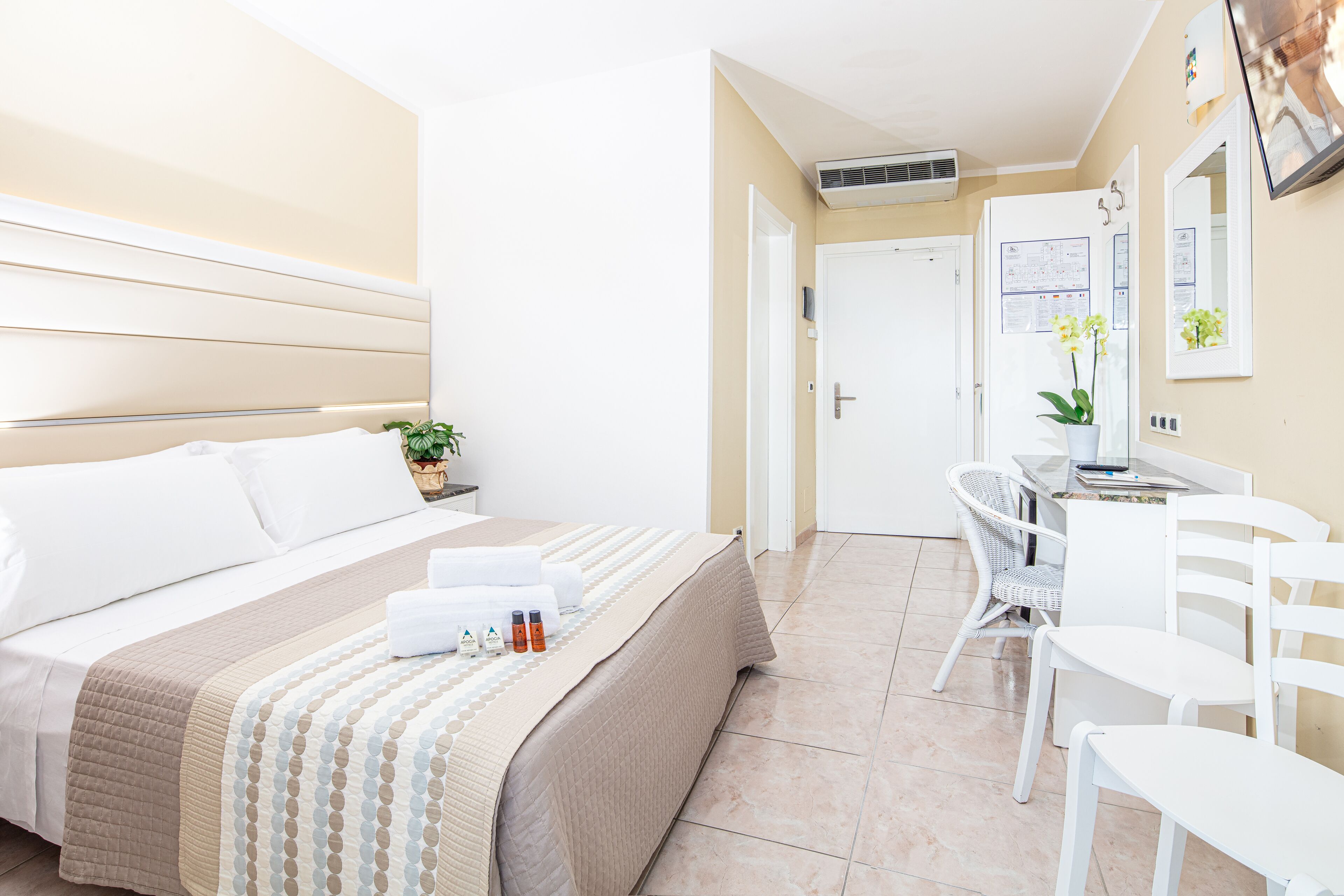 Quarto de hotel com cama, escrivaninha e cadeira. Há uma planta na escrivaninha e uma porta ao fundo.
