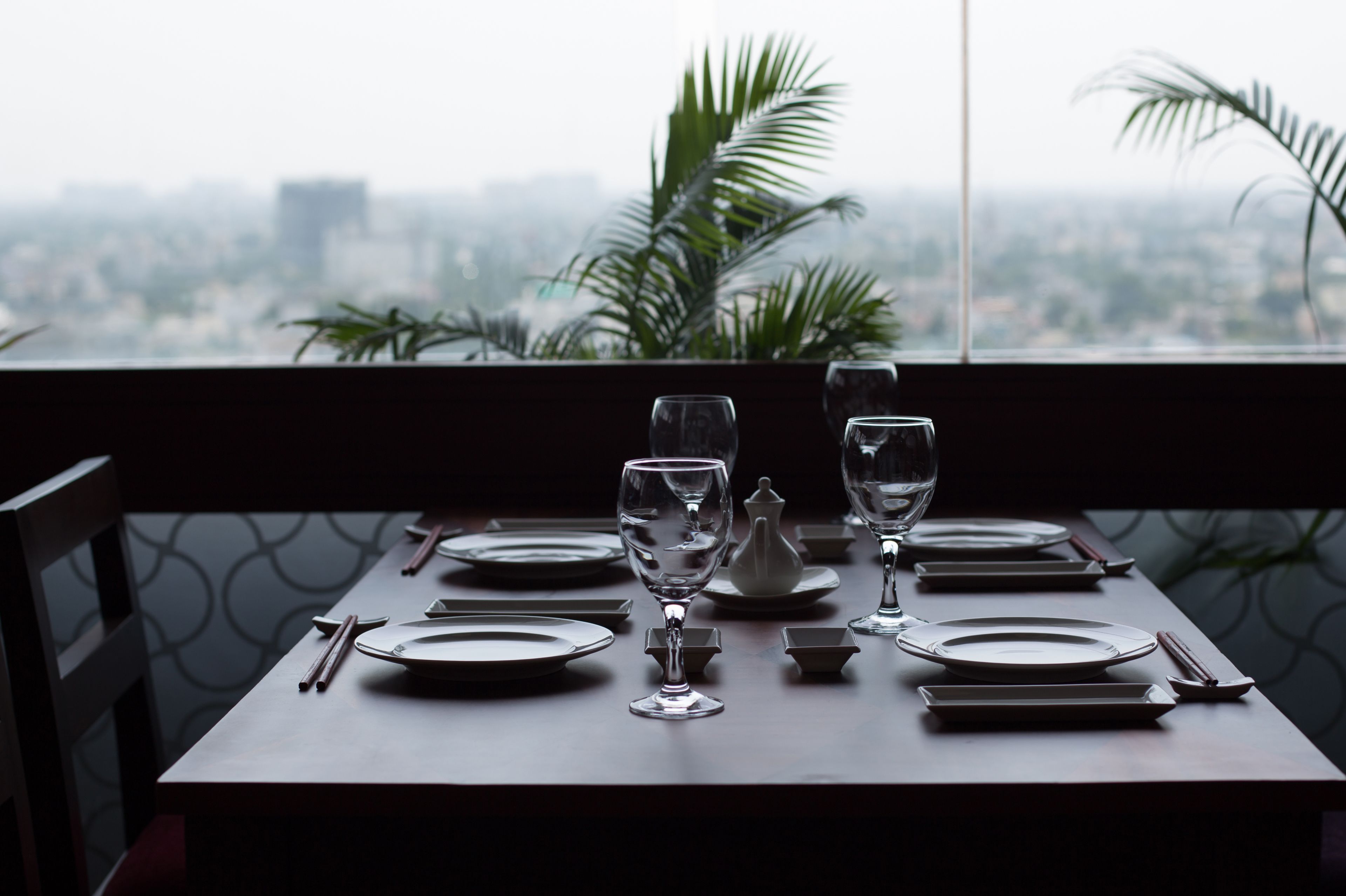 Uma mesa de jantar posta com pratos, talheres e copos, com vista para a cidade através de uma janela.