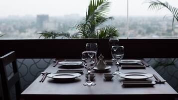 Uma mesa de jantar posta com pratos, talheres e copos, com vista para a cidade através de uma janela.