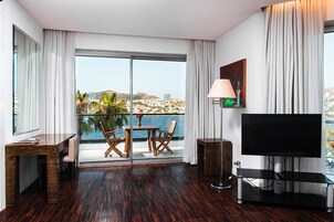 Quarto de hotel moderno com varanda, televisão de tela plana, mesa de jantar de madeira e vista para a cidade e mar.