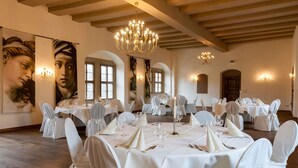 Uma sala de banquetes com mesas redondas preparadas para refeições, cadeiras brancas, lustres e grandes pinturas nas paredes.