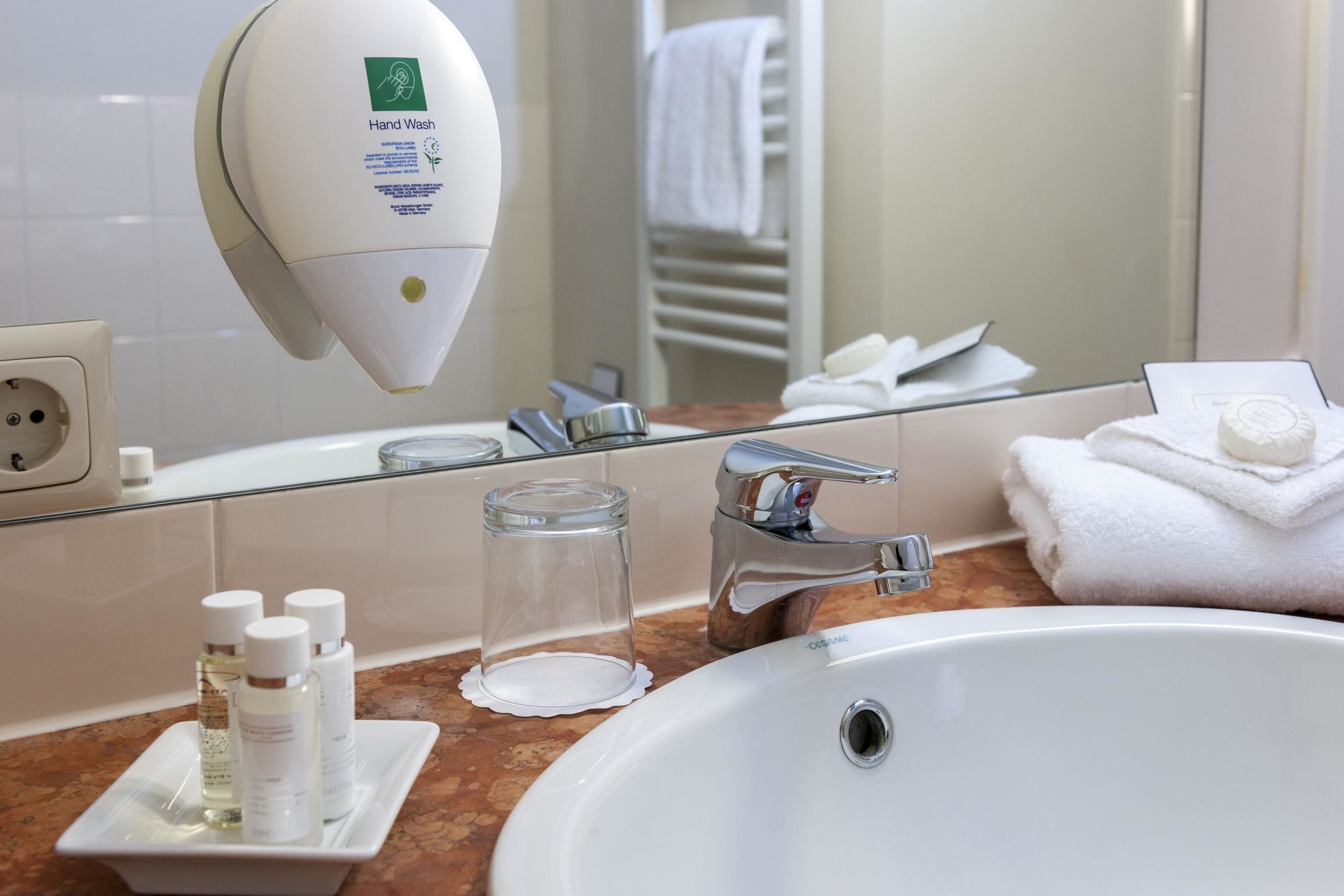 Casa de banho de hotel com dispensador de sabonete líquido, pia e artigos de higiene pessoal no balcão.