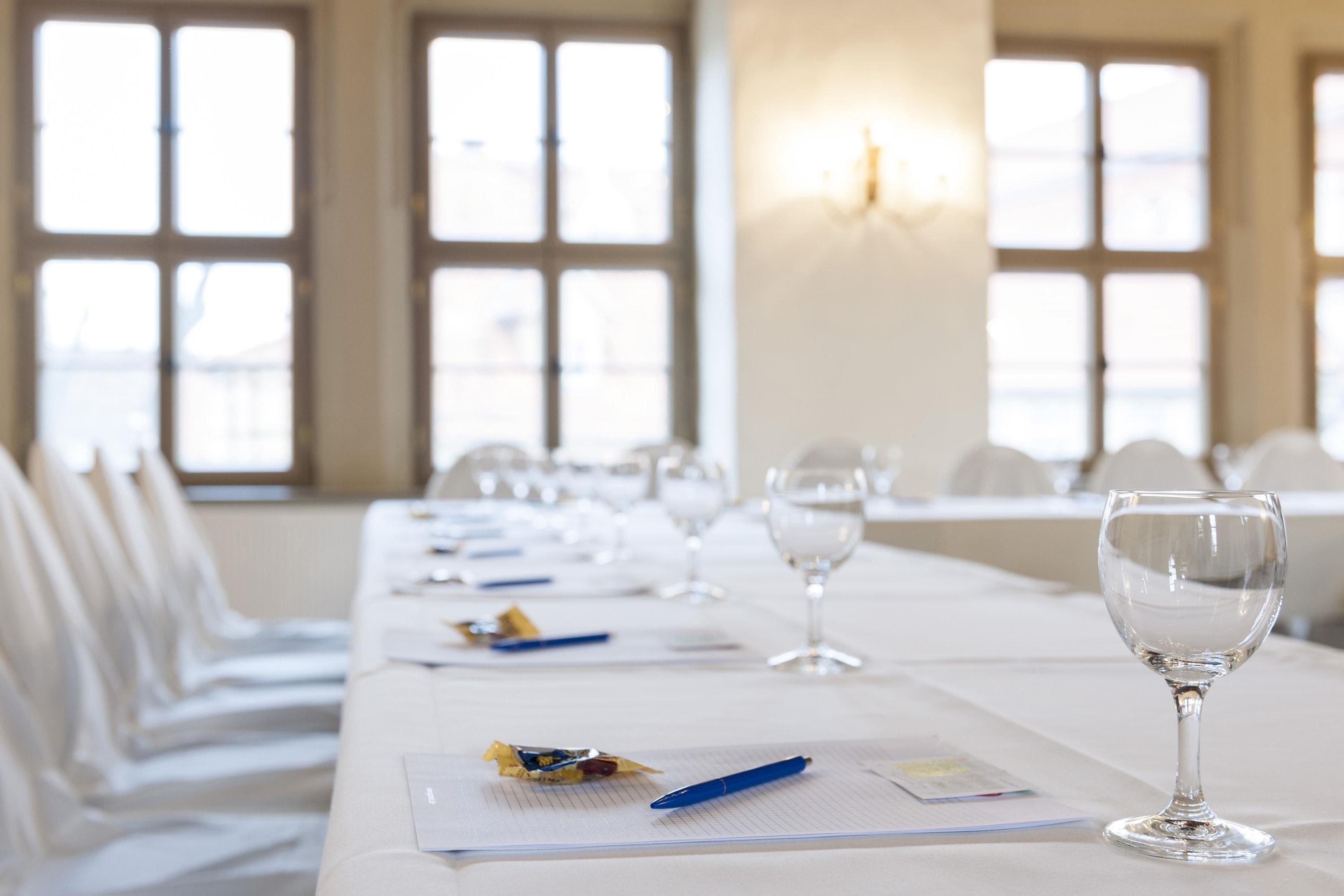 Uma mesa comprida com toalhas brancas, disposta com cadeiras e copos de vidro transparentes.