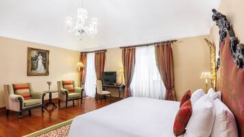 Quarto de hotel com uma cama grande, duas poltronas, uma escrivaninha com computador, televisão e um candelabro.