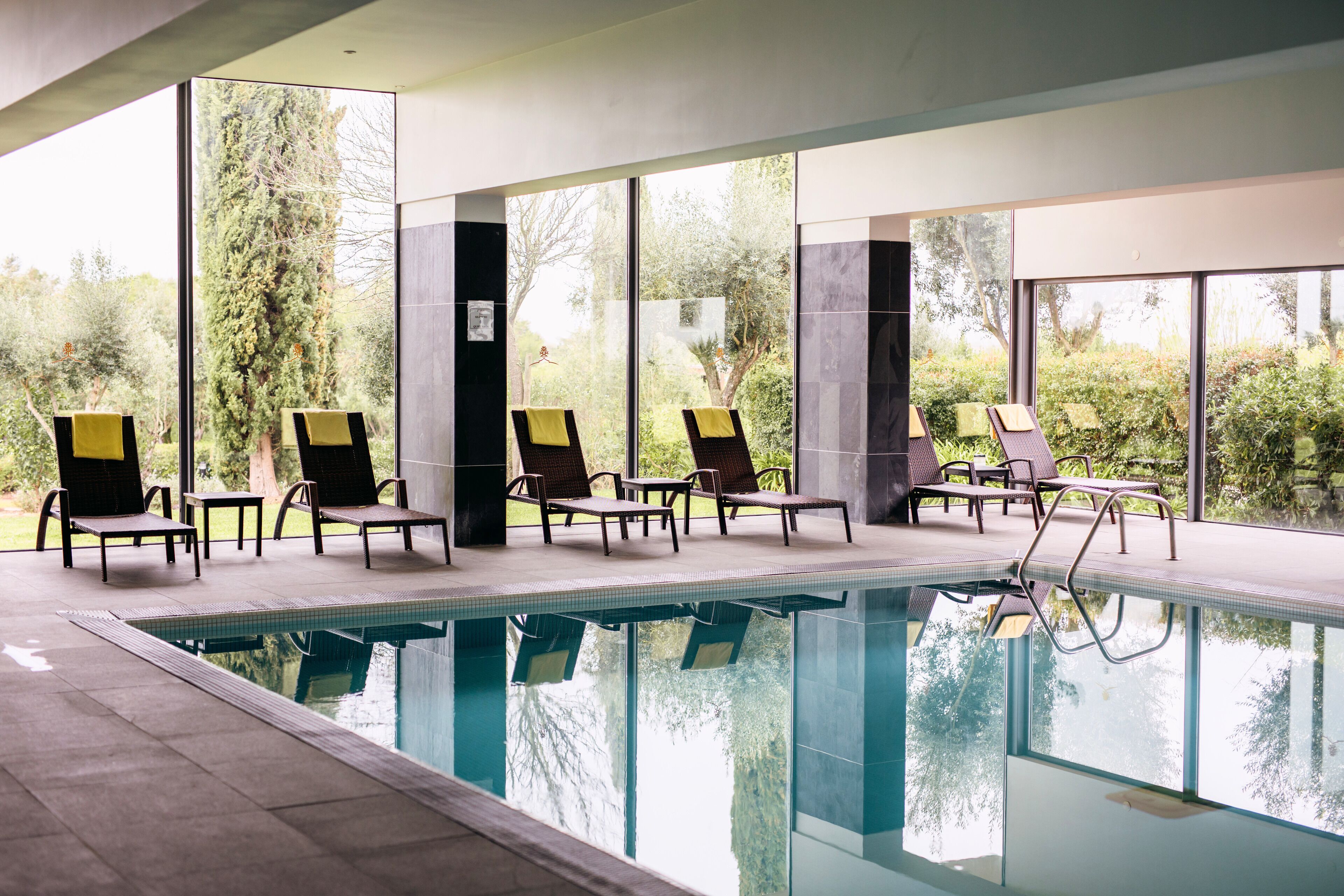 Piscina interior com paredes de vidro transparente, cercada por espreguiçadeiras e mesas, com vista para o verde lá fora.