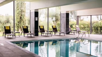 Piscina interior com paredes de vidro transparente, cercada por espreguiçadeiras e mesas, com vista para o verde lá fora.