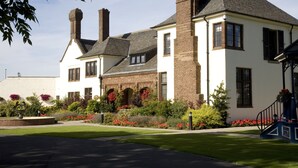 Uma casa grande de dois andares com chaminé de tijolos, paredes brancas e um jardim bem cuidado com flores vermelhas.