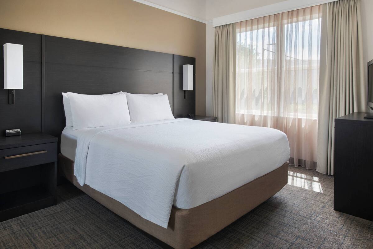 Quarto de hotel com uma cama grande, mesa de cabeceira, televisão e janela com cortinas.