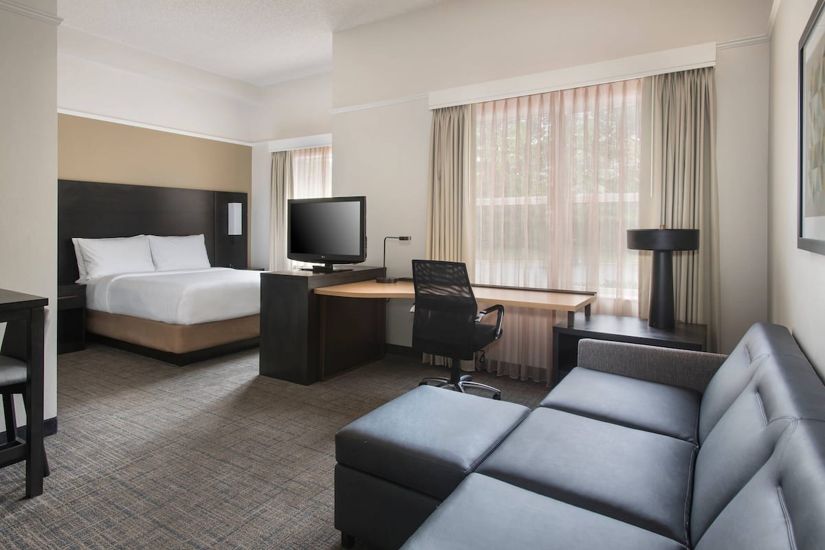 Quarto de hotel com cama, escrivaninha com computador, televisão, sofá e janela com cortinas.