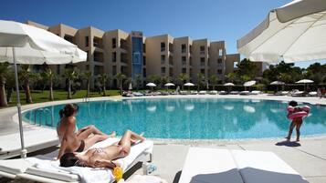 Área de piscina com espreguiçadeiras, guarda-sóis e um edifício ao fundo.
