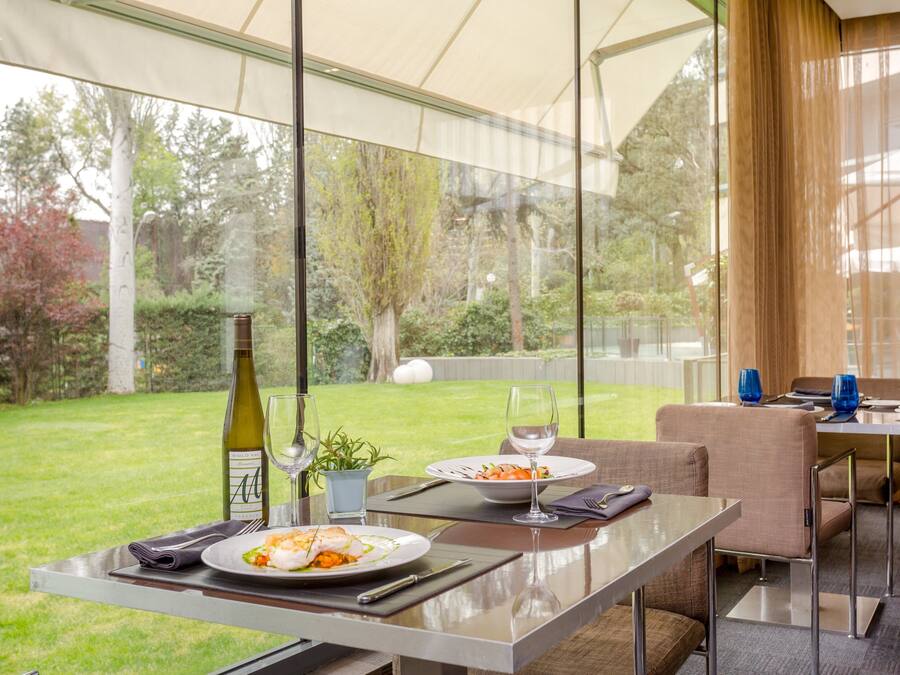Uma área de refeições com uma mesa posta para dois, uma garrafa de vinho e vista para um jardim através de grandes janelas.