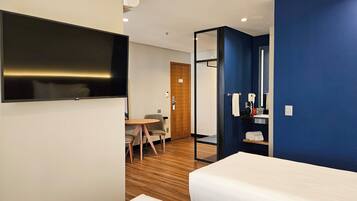 Quarto de hotel com cama, escrivaninha, cadeira, televisão e banheiro com espelho e porta-toalhas.