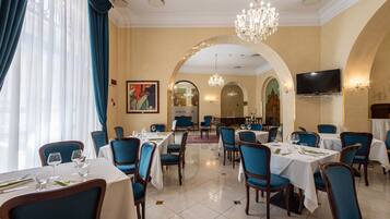 Uma sala de jantar formal com mesas postas, cadeiras estofadas azuis e um candelabro.
