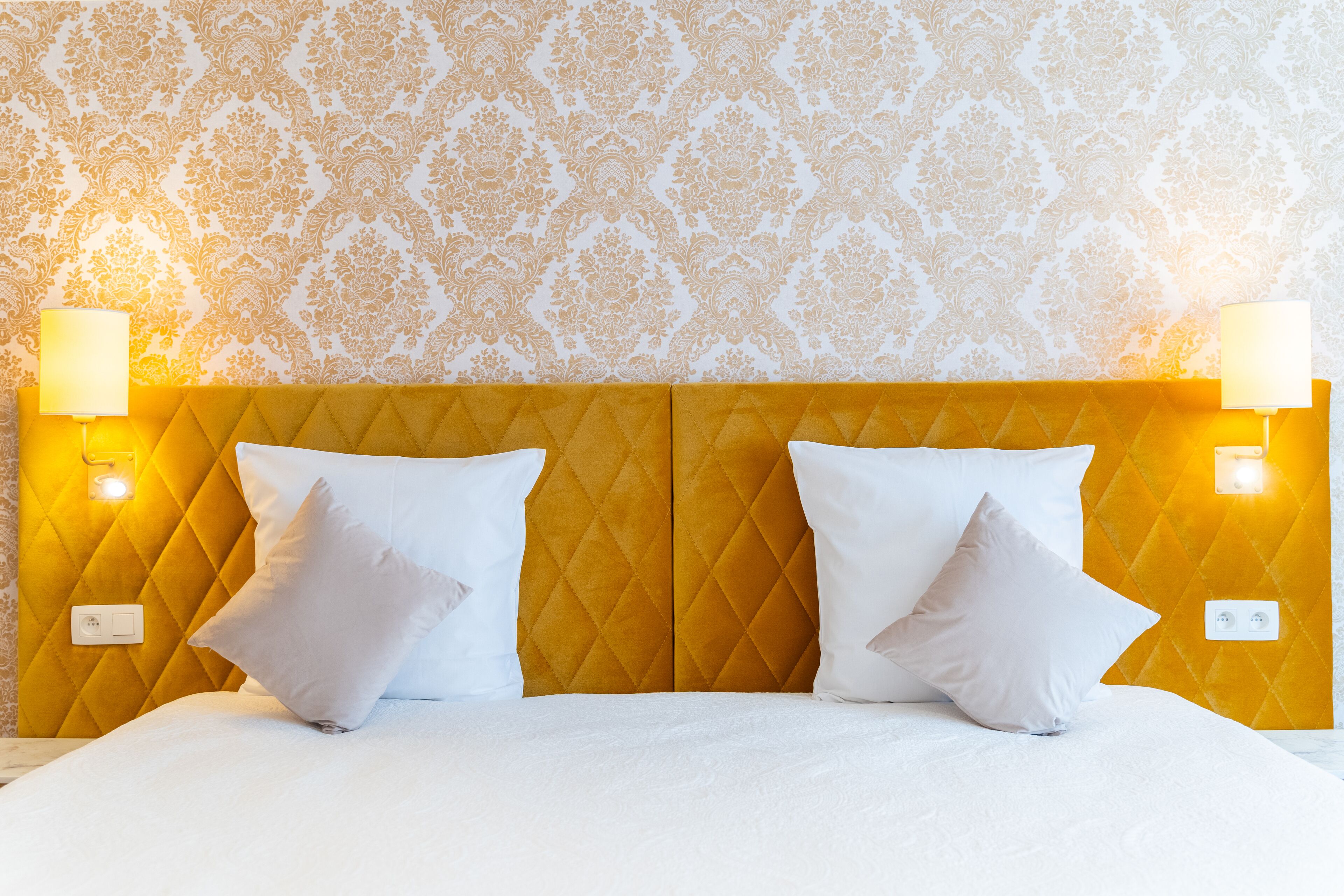Quarto de hotel com cabeceira acolchoada, duas almofadas brancas e luminárias fixadas na parede.