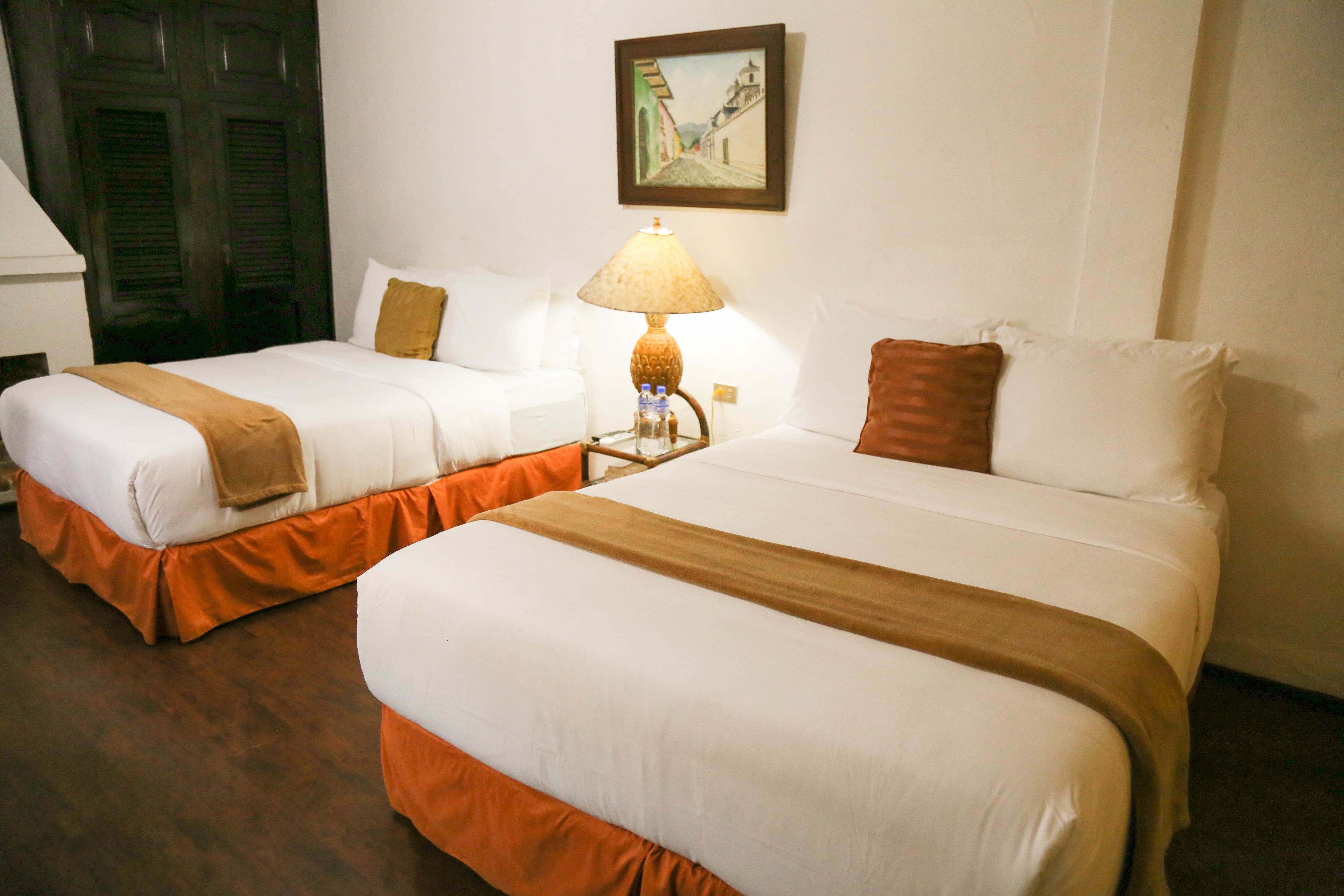 Quarto de hotel com duas camas, uma mesa de cabeceira com luminária e um quadro na parede.