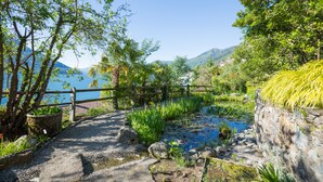 Um jardim tranquilo com lago, caminho de pedra e cerca de madeira, com vista para um corpo de água e montanhas.