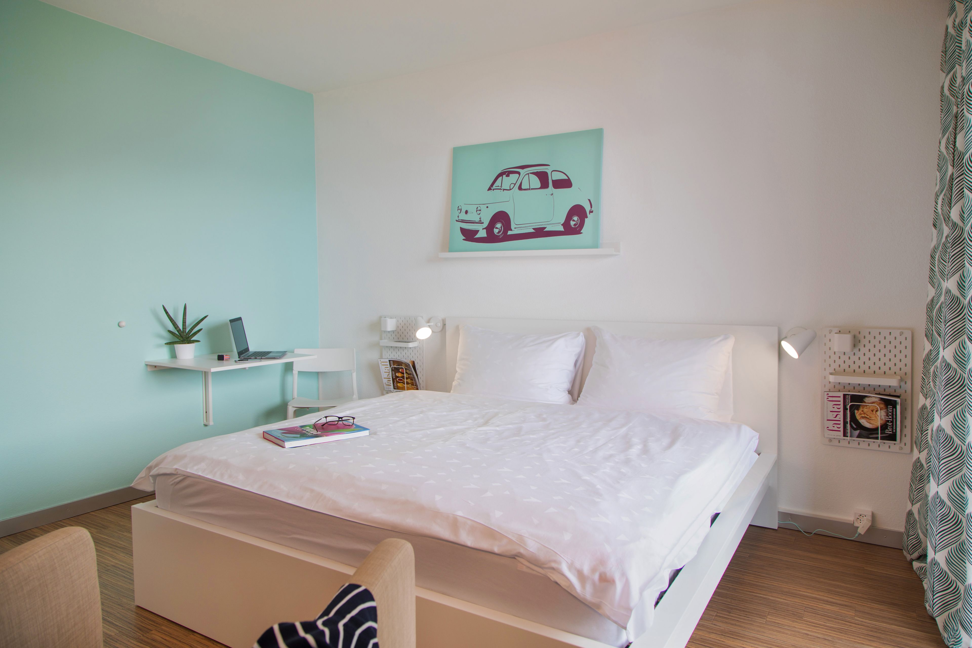 Um quarto com cama, uma escrivaninha com um laptop, uma pequena planta e um prateleira fixada na parede.