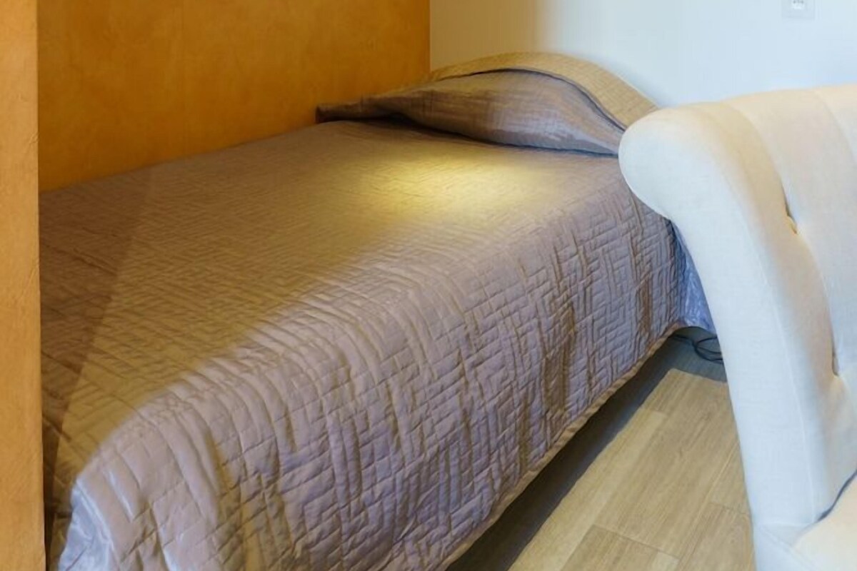 Quarto pequeno com cama de solteiro, uma cama simples, uma cadeira branca e um interruptor na parede.