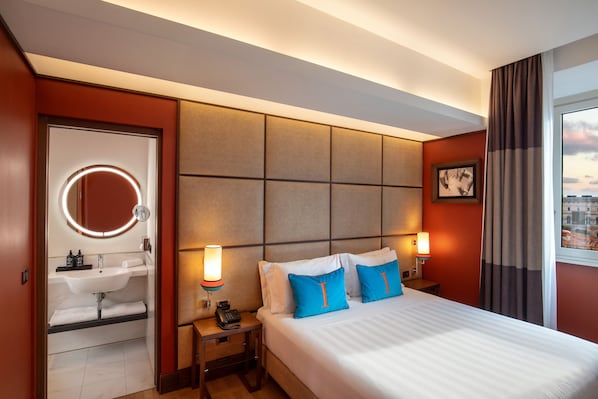Quarto de hotel com cama, mesas de cabeceira, um espelho redondo, banheiro com pia e vista para a cidade pela janela.