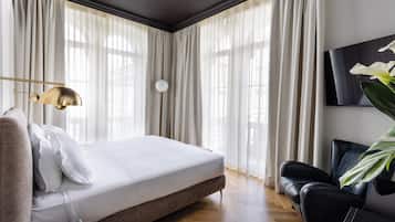 Quarto moderno com cama, poltrona de couro preto, televisão na parede e cortinas translúcidas.
