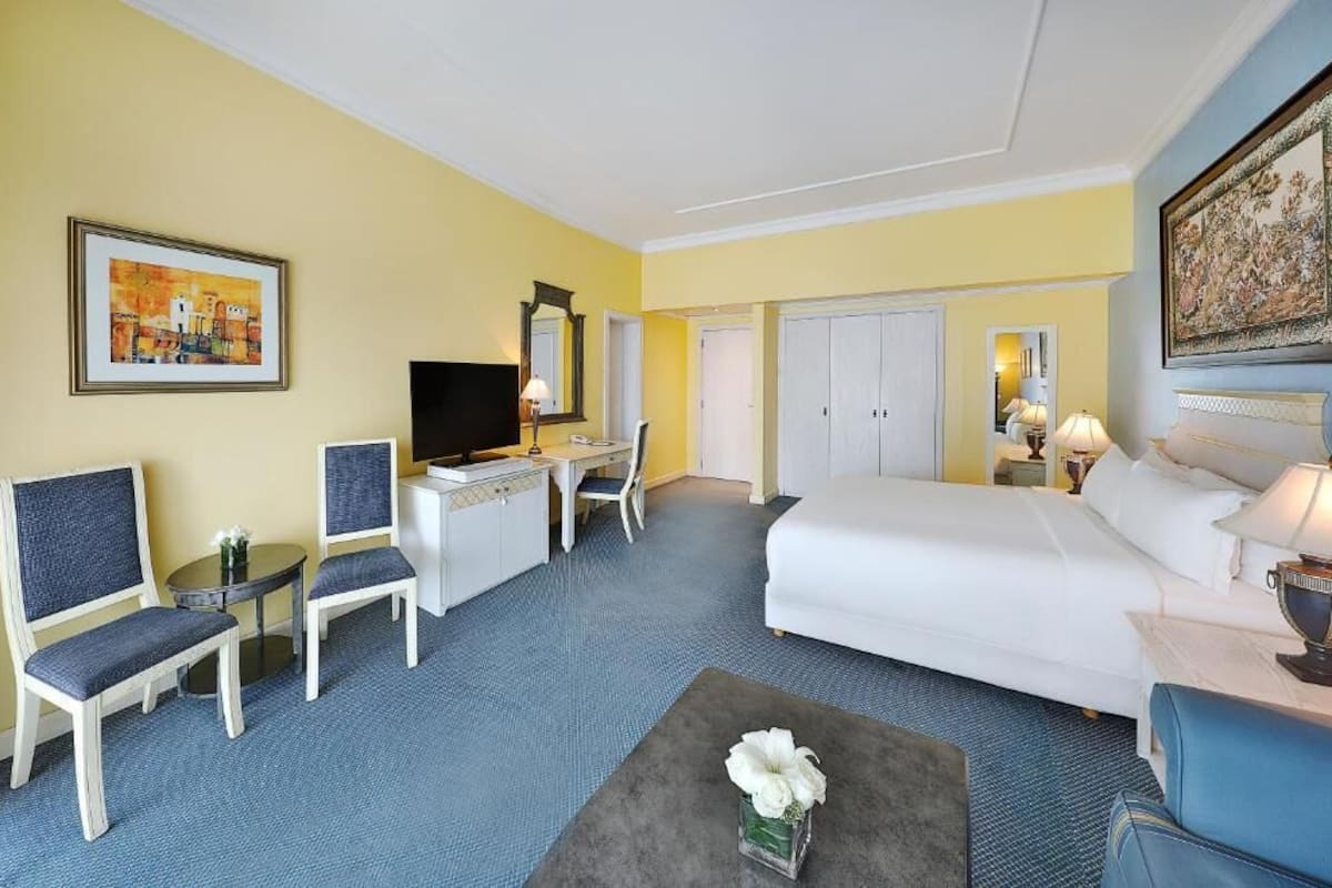 Quarto de hotel com cama, escrivaninha, televisão e duas cadeiras.