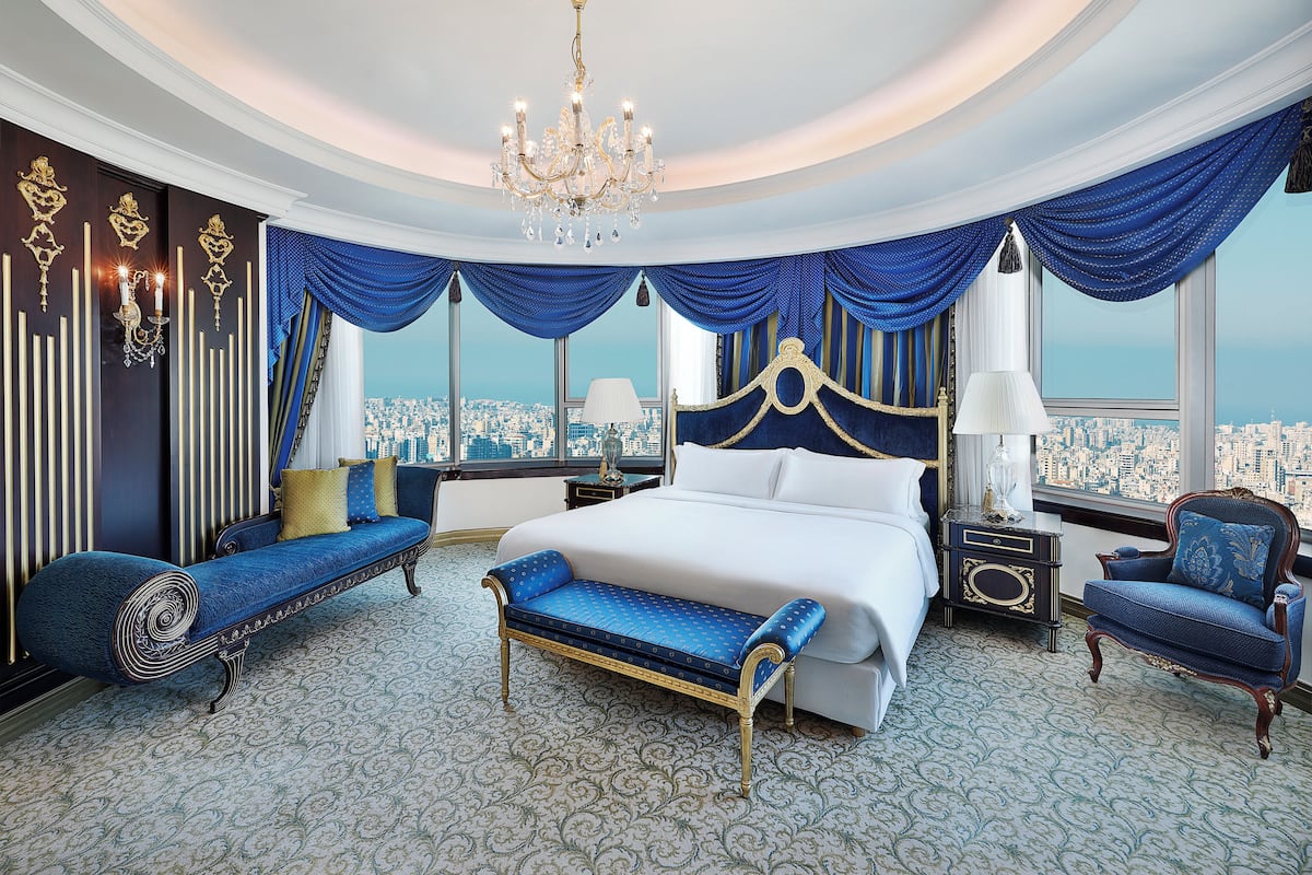 Quarto luxuoso com uma cama grande, um banquinho e uma cadeira, oferecendo vista da cidade por grandes janelas.