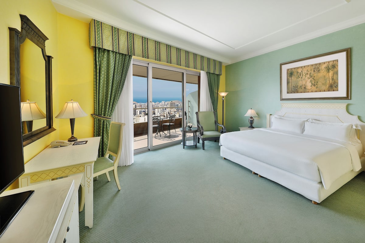 Quarto de hotel com cama, escrivaninha, cadeira, varanda com vista para a cidade e um quadro na parede.