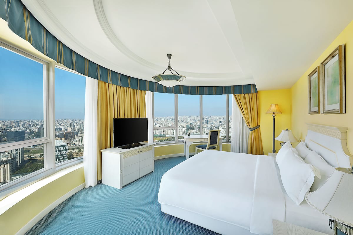Quarto de hotel com uma cama grande, uma mesa, uma televisão e vista para a cidade.