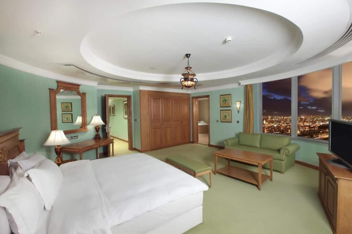 Quarto de hotel com cama, sofá, televisão e uma grande janela vista para a cidade.