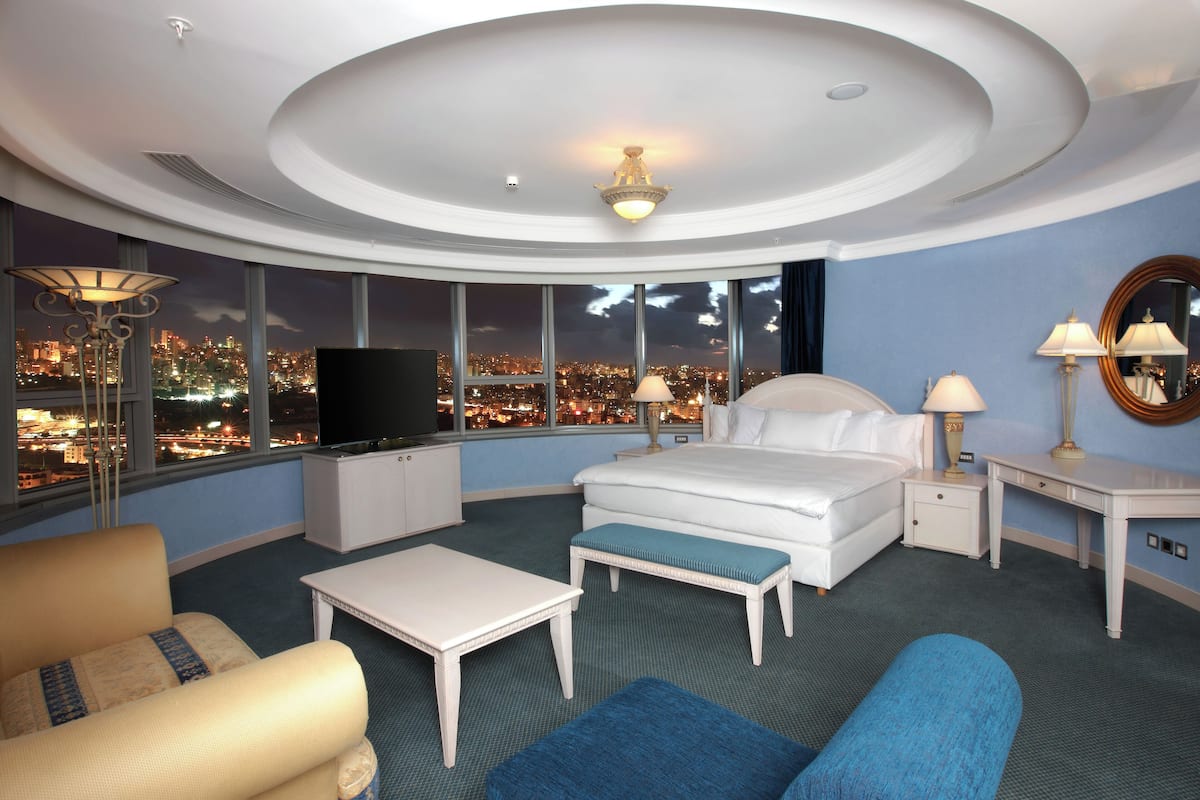 Quarto de hotel com cama, escrivaninha, cadeira, televisão e vista para a cidade à noite.