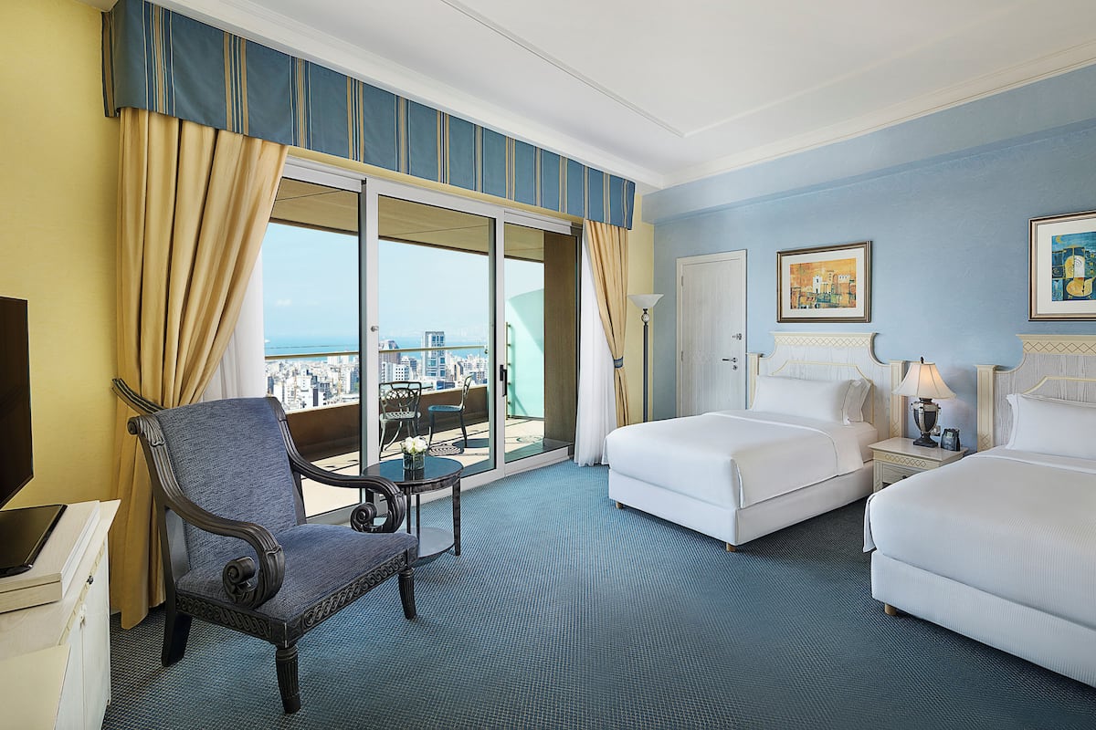 Quarto de hotel com varanda, uma cama, escrivaninha e cadeira, e vista para a cidade.