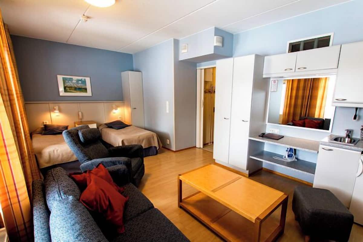 Quarto de hotel compacto com sofá, duas camas de solteiro, uma kitchenette pequena e um espaço para refeições.