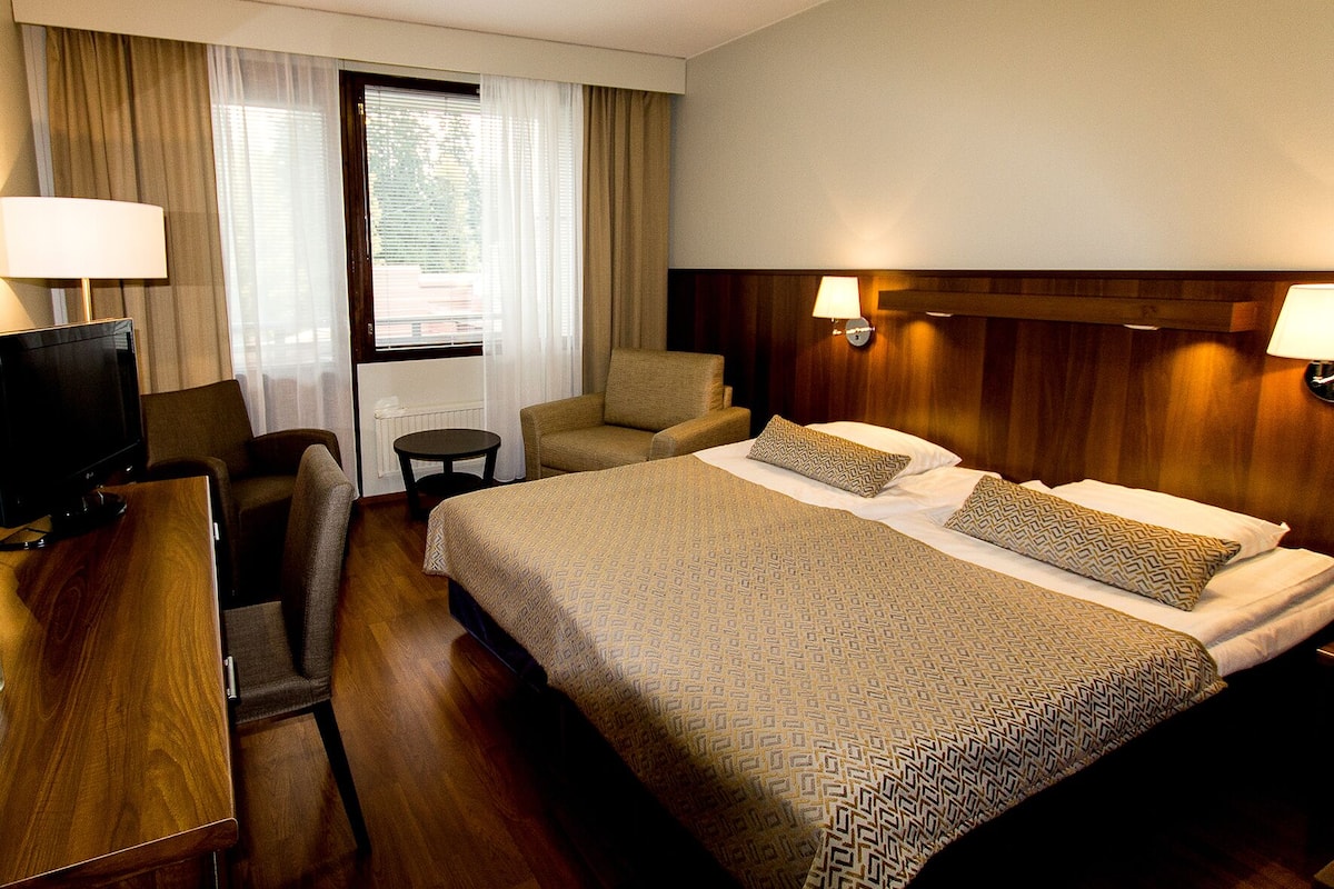 Quarto de hotel com uma cama grande, duas poltronas, uma escrivaninha com televisão e uma janela com cortinas.