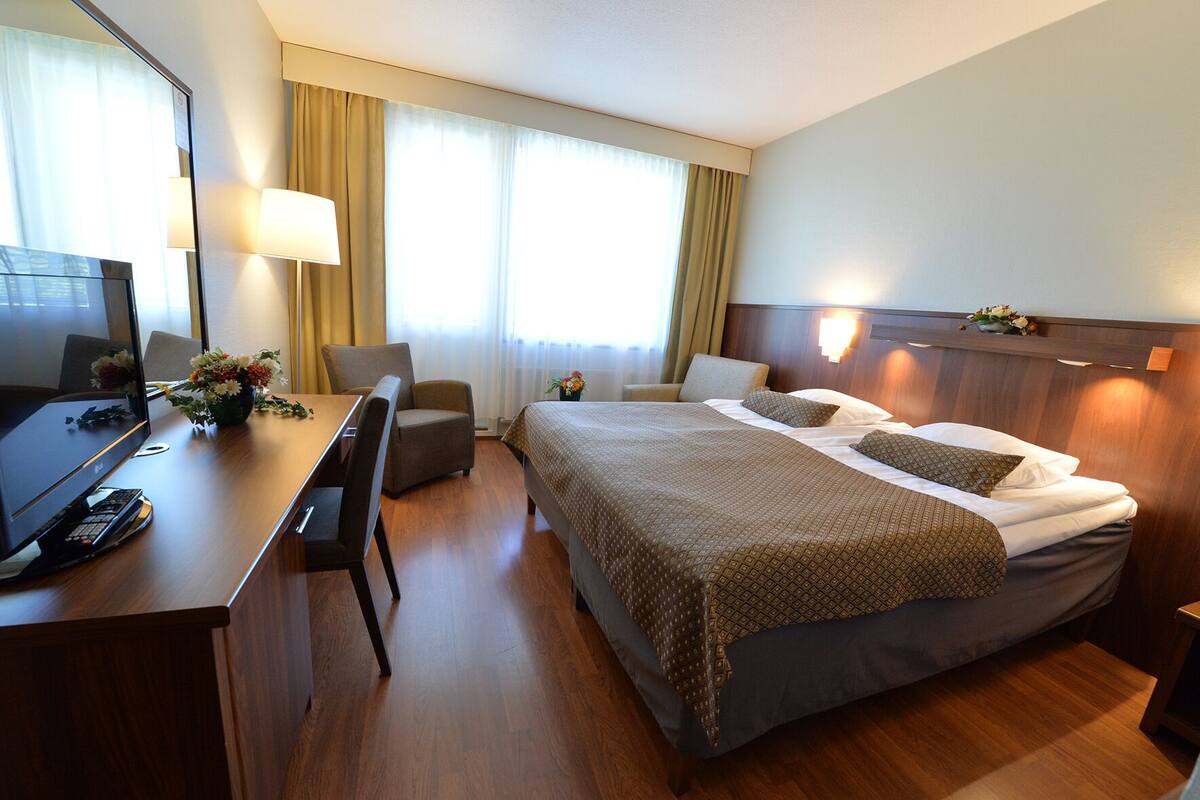 Quarto de hotel com cama, escrivaninha, cadeira, televisão e janela com cortinas.