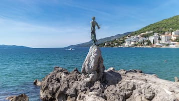 Uma estátua em um afloramento rochoso à beira-mar, com edifícios ao fundo.