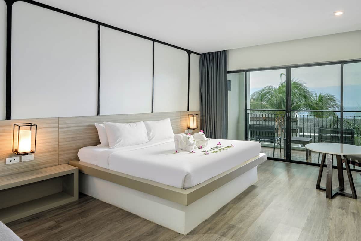 Quarto de hotel moderno com uma cama grande, luminárias de cabeceira, uma mesinha, uma cadeira e uma TV fixada na parede.