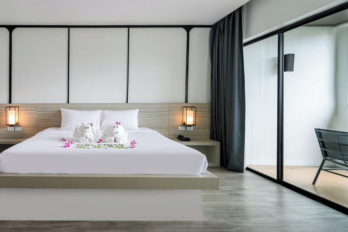Quarto de hotel moderno com uma cama grande, duas mesas de cabeceira com luminárias, um banco ao pé da cama e vista para o exterior através de uma porta de vidro.