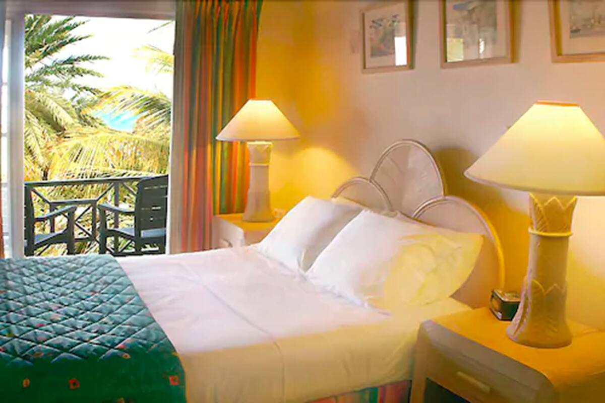 Quarto de hotel com cama, duas luminárias de cabeceira, mesa de apoio e vista para palmeiras pela janela.