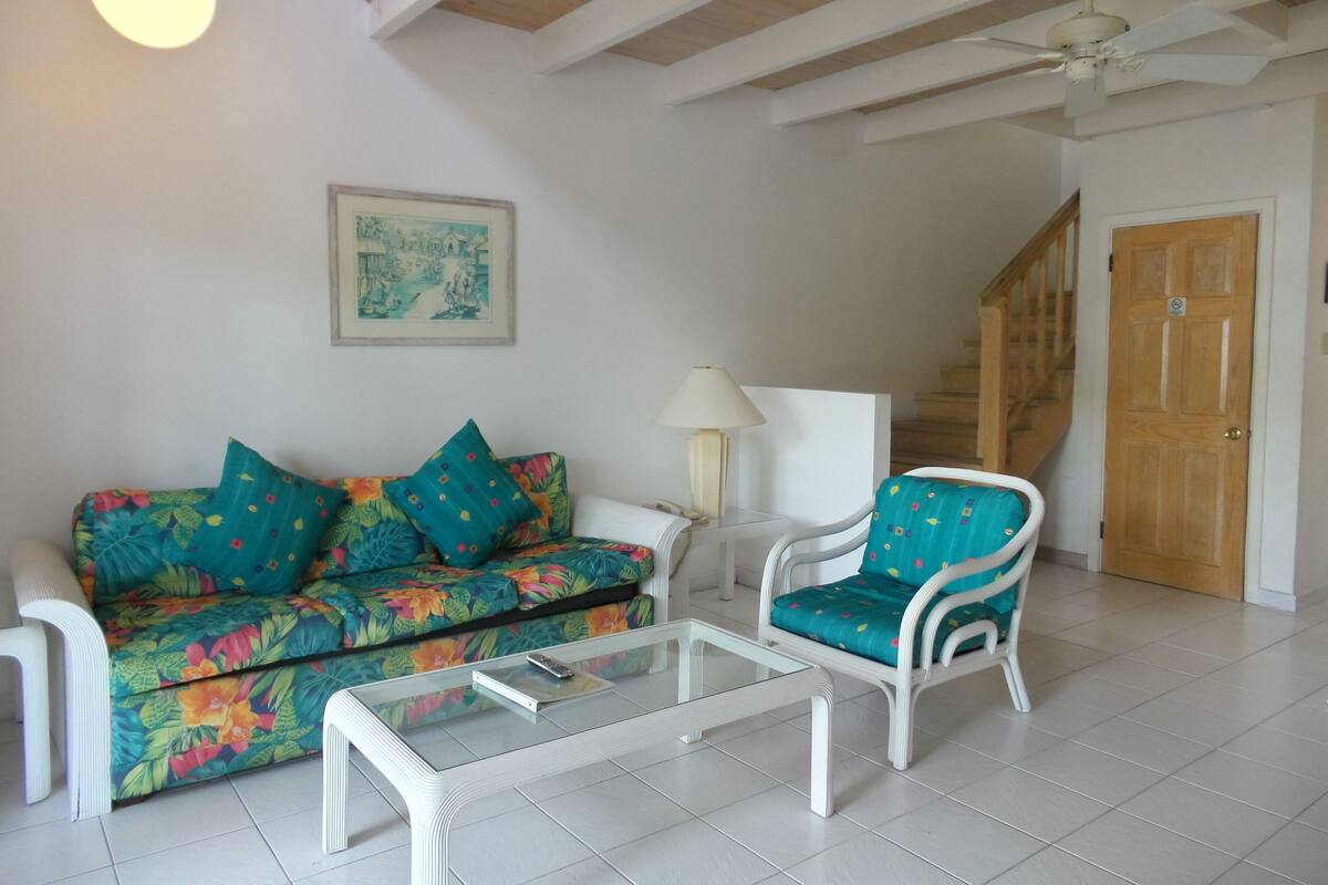 Sala de estar com sofá, poltrona e mesa de centro. Escada e porta ao fundo.