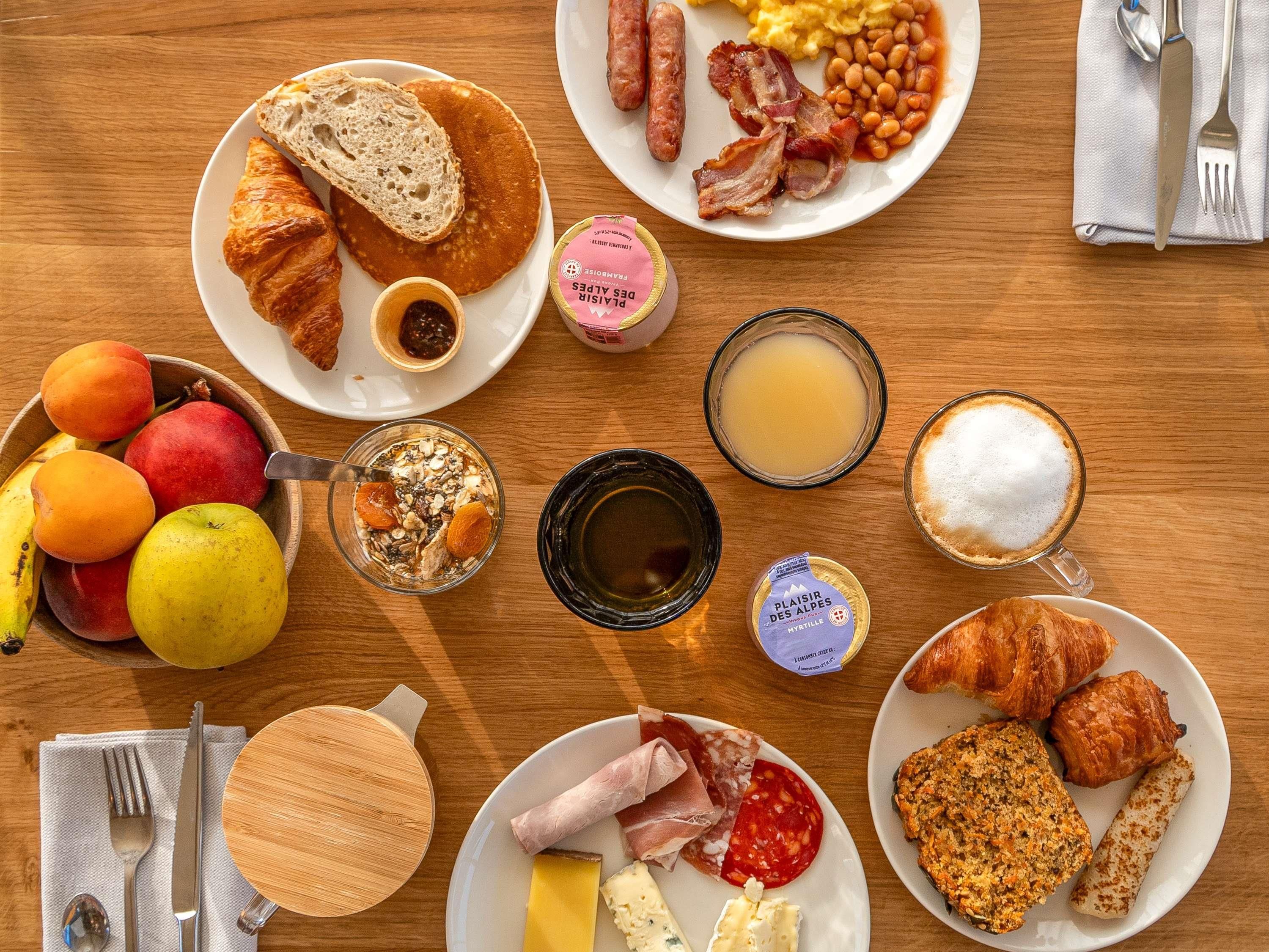 Mesa de café da manhã com croissants, pão, linguiça, bacon, feijão, ovos, torradas, frutas, geleia, café e queijo.