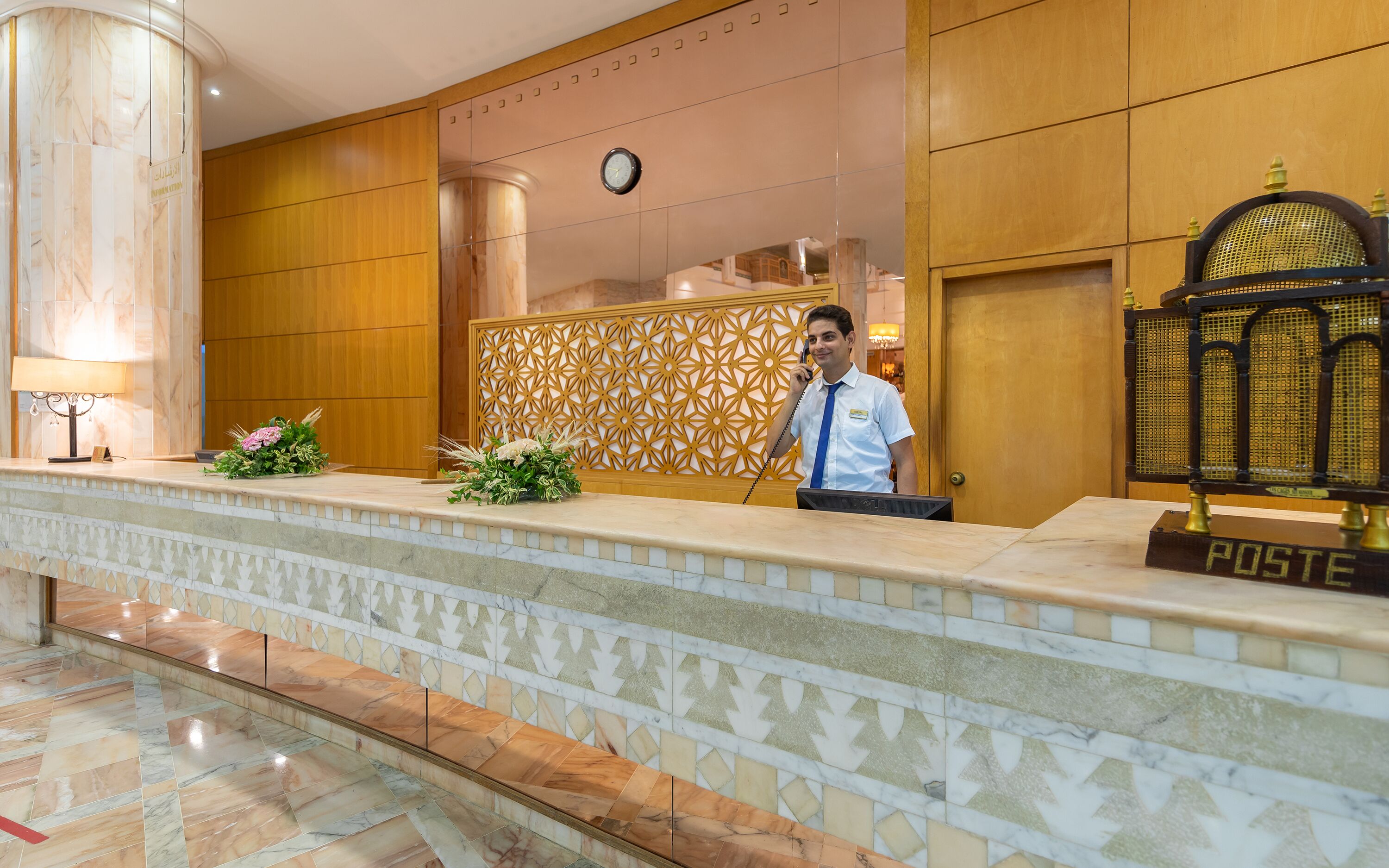 Bancada de recepção de hotel com painel de madeira ao fundo, um relógio e um ecrã decorativo.