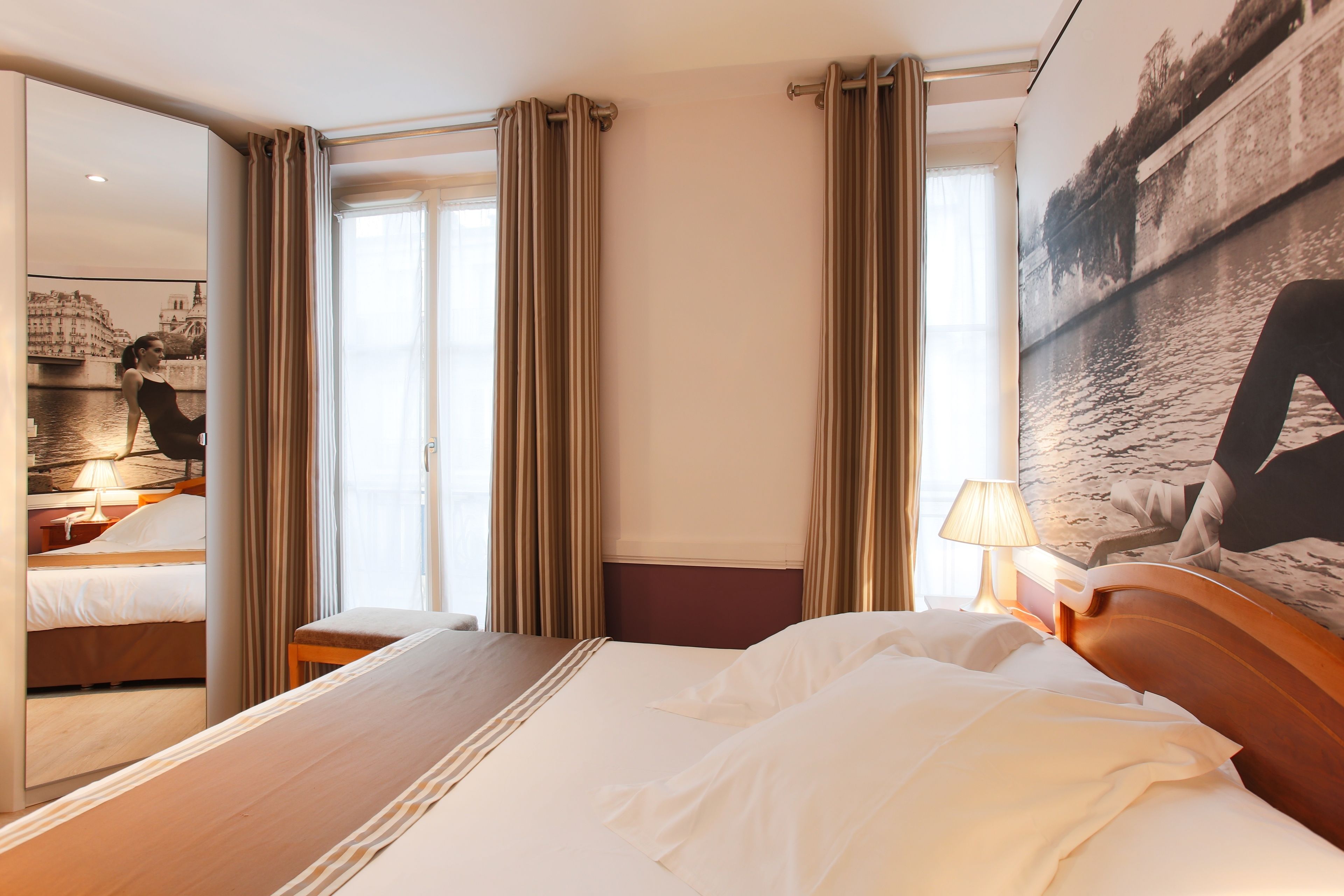 Quarto de hotel com uma cama grande, mesa de cabeceira com abajur, espelho e vista para um rio pela janela.