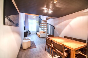 Um interior moderno com mesa de jantar de madeira, cadeiras, escada em espiral e um sofá em uma sala de estar iluminada com grandes janelas.