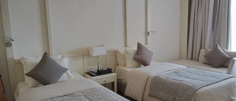 Quarto de hotel com duas camas, uma mesinha de cabeceira com abajur e uma janela com cortinas.