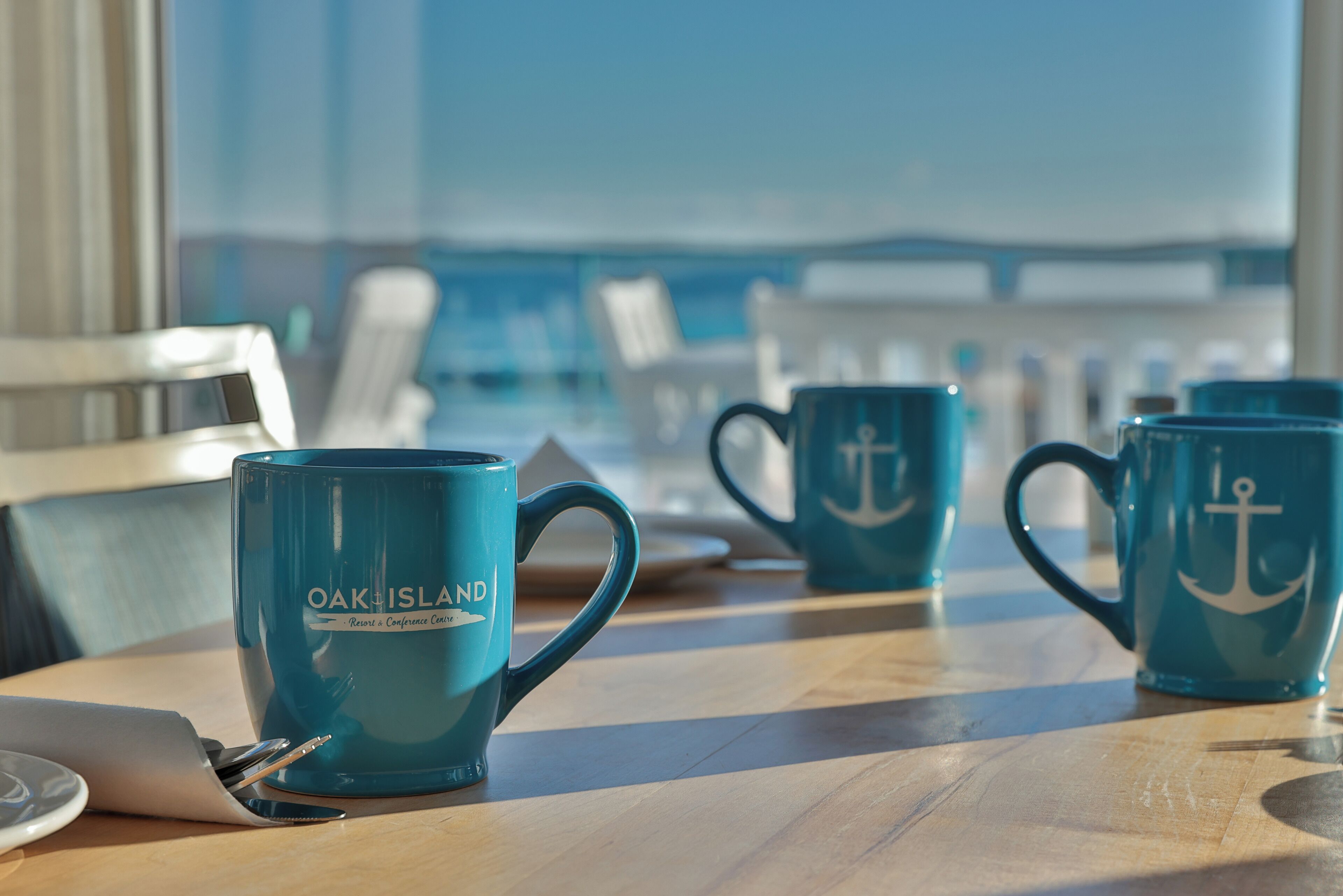 Uma mesa com quatro canecas verde-água com a inscrição "Oak Island" e um logotipo de âncora, sobre uma mesa de madeira com vista para cadeiras brancas e um corrimão ao fundo.